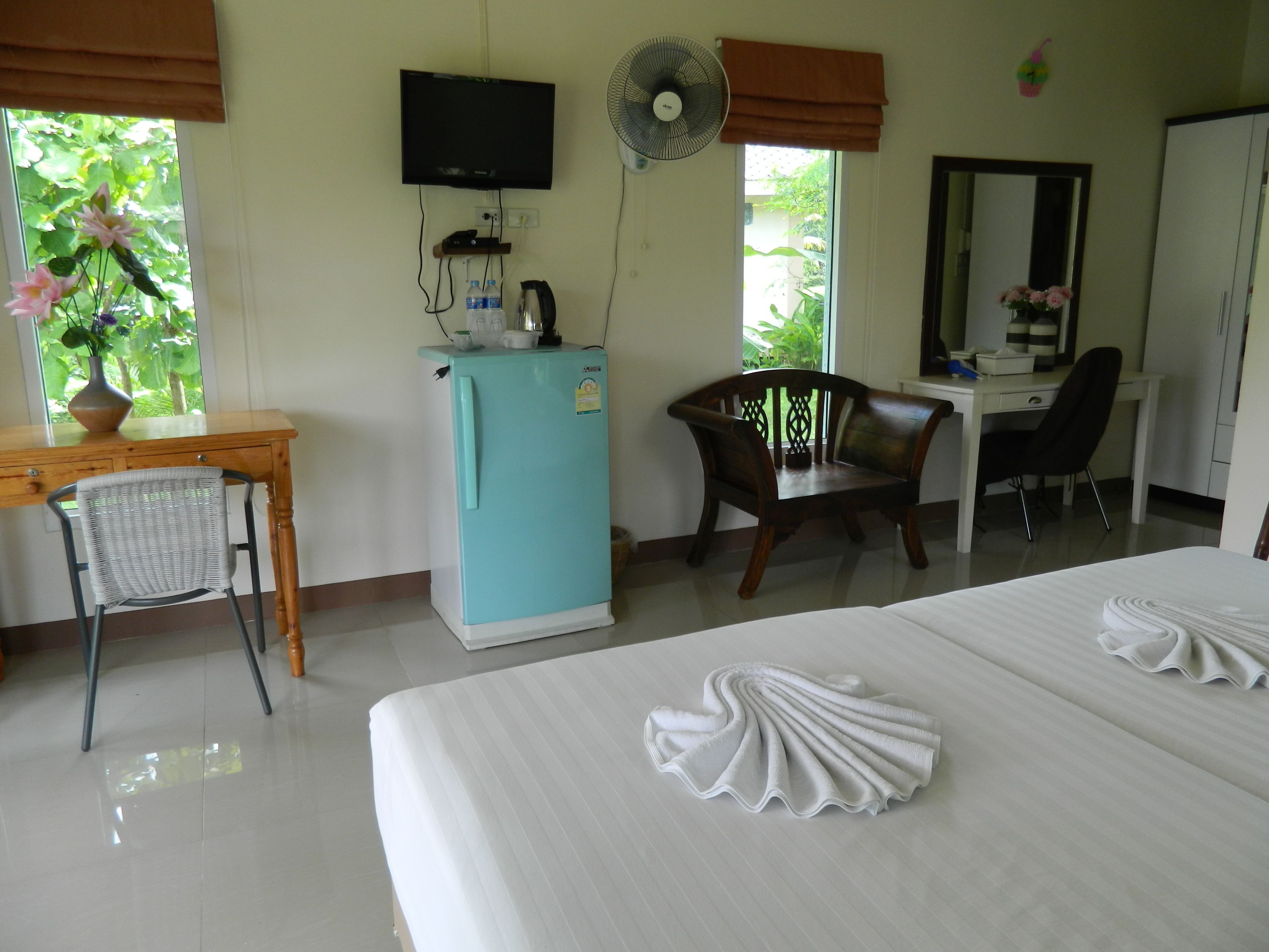 Quarto de hotel com cama, escrivaninha com cadeira, mini-frigorífico, televisão, ventoinha e janela com vista para área verde.