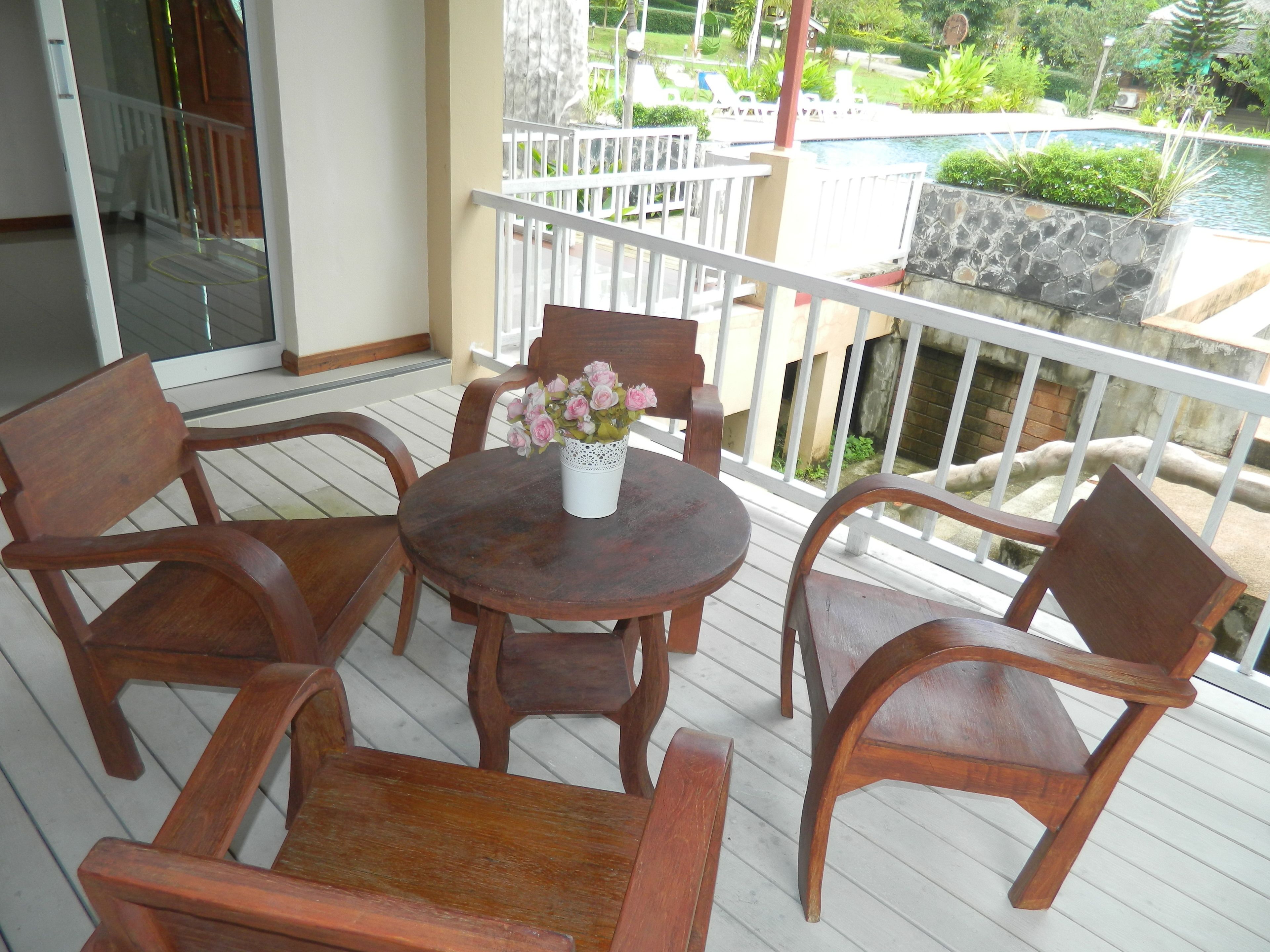Um deck de madeira com uma mesa redonda e cadeiras, guarda-corpo branco e vista para uma piscina e vegetação.