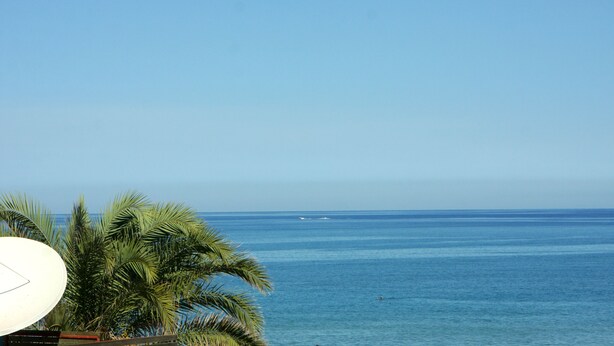 Visão nítida do oceano, com um barco ao longe, palmeiras e um guarda-sol branco.