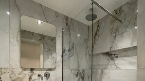 Um banheiro moderno com parede de mármore, toucador de madeira, pia branca, box de vidro e um espelho.