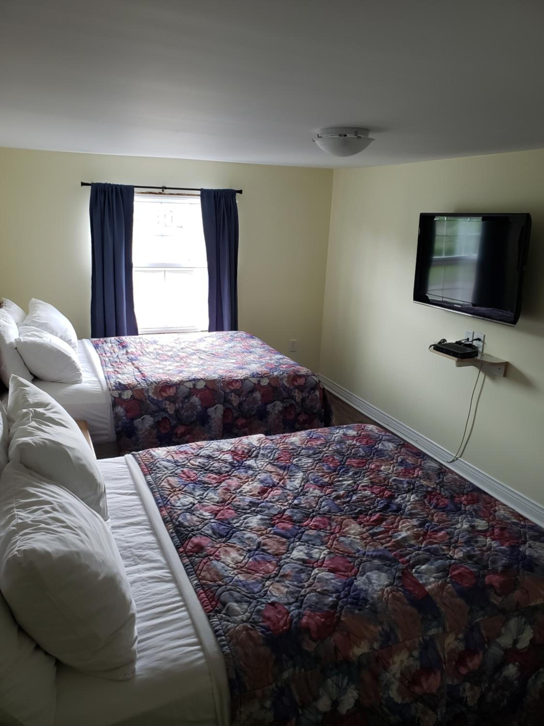 Quarto com duas camas, televisão fixada na parede e janela com cortinas.