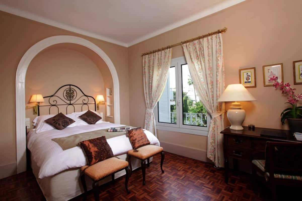 Um quarto com uma cama, duas luminárias de cabeceira, uma cadeira, uma escrivaninha e uma janela com cortinas.