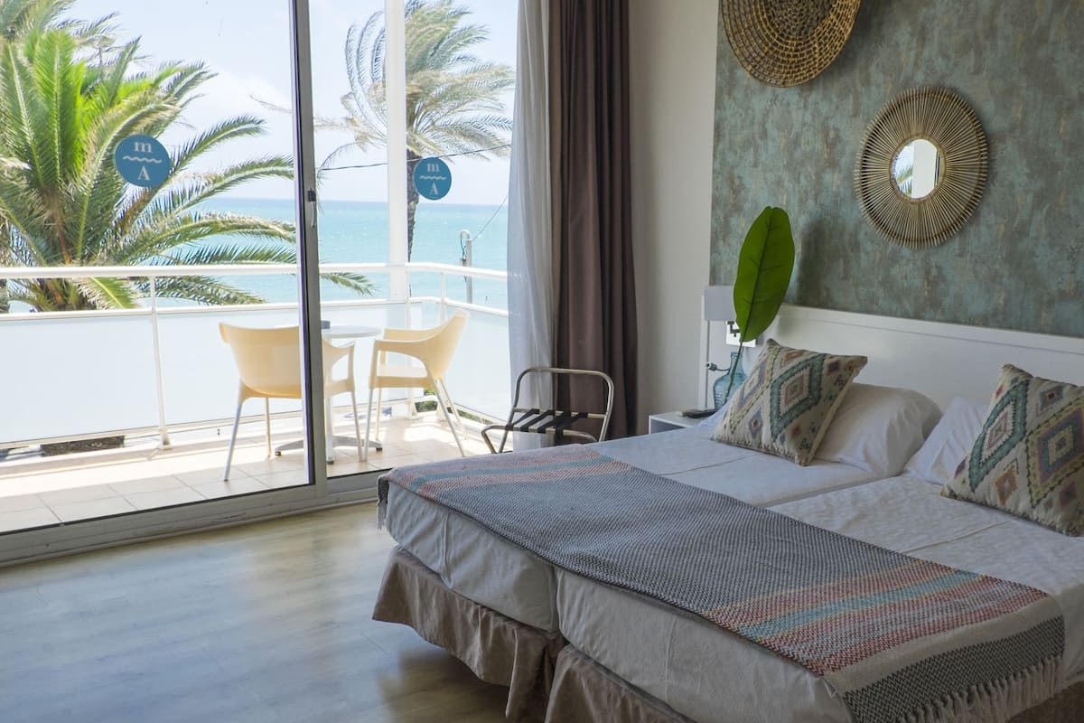 Quarto de hotel com uma cama grande, mesa de cabeceira com telefone, cadeira e vista para o oceano, além de varanda com cadeiras.