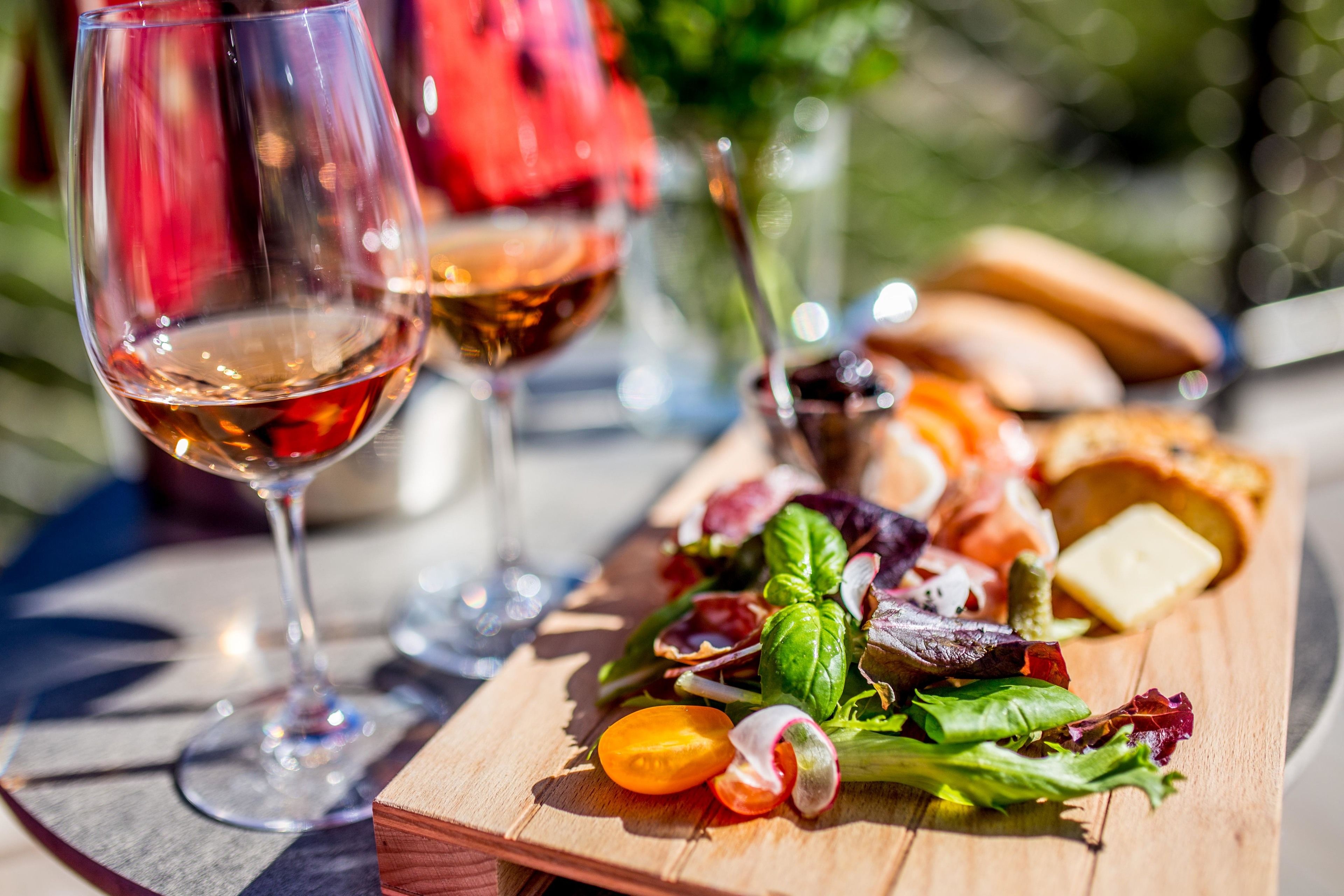 Uma tábua de madeira com diversos queijos, carnes e vegetais, acompanhada por duas taças de vinho tinto sobre uma mesa ao ar livre.