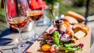 Uma tábua de madeira com diversos queijos, carnes e vegetais, acompanhada por duas taças de vinho tinto sobre uma mesa ao ar livre.