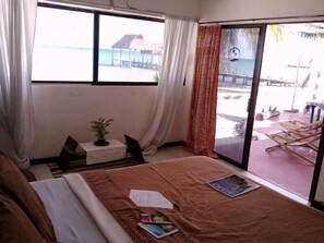 Quarto com cama, uma mesinha com uma planta e vista para uma praia através de uma porta de correr.
