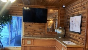 Interior de cabana de madeira com teto de madeira, TV fixada na parede, lareira de pedra e janela com vista.