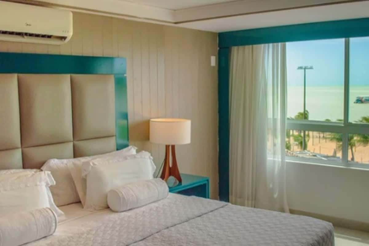 Quarto de hotel com uma cama, mesa de cabeceira, abajur e vista para a praia.
