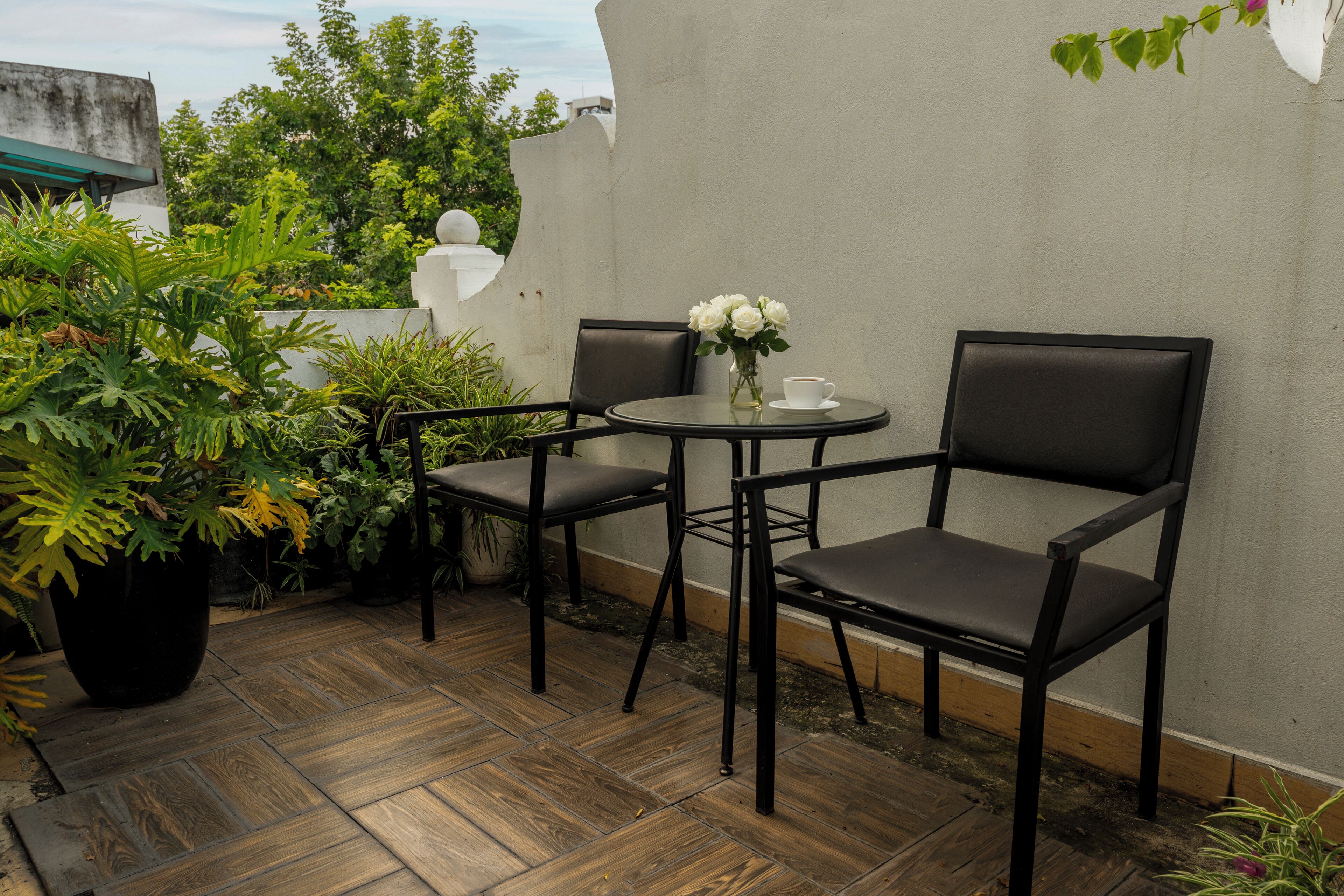 Uma área de pátio com uma mesa redonda e duas cadeiras, cercada por plantas em vasos e uma parede branca.