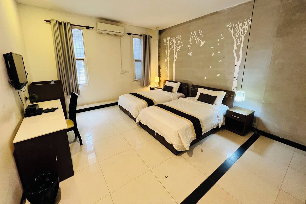 Quarto de hotel com duas camas, uma escrivaninha, uma televisão e um mural de uma árvore na parede.