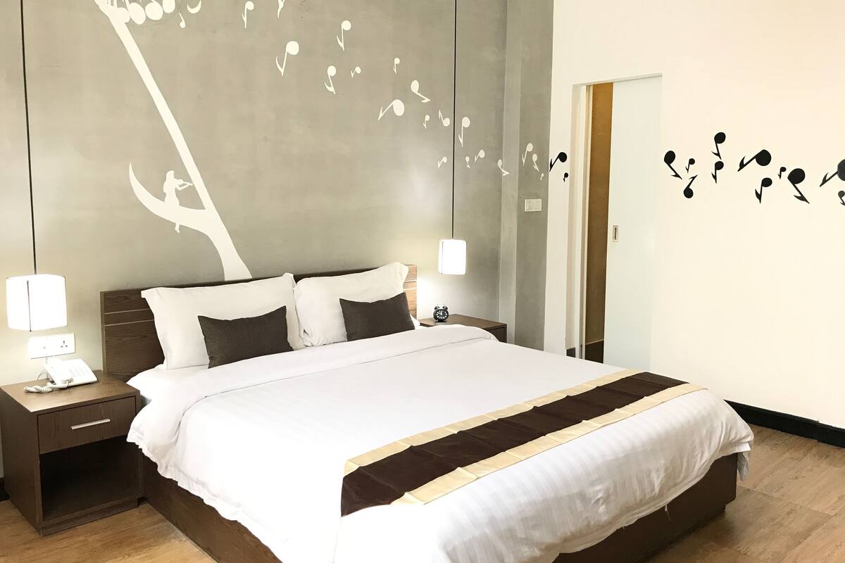 Quarto de hotel moderno com uma cama grande, duas mesas de cabeceira com abajures, um mural na parede e uma porta.