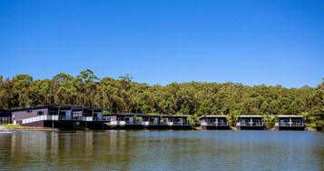 Fileira de cabanas modernas à beira-mar, com várias andares e varandas, com a densa floresta e céu azul límpido ao fundo.