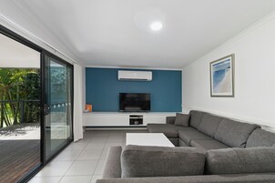 Uma sala de estar moderna com um sofá seccional cinza, uma TV de tela plana em um móvel branco e uma grande porta de vidro deslizante que dá para um deck externo.