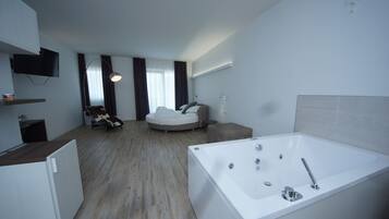 Quarto de hotel moderno com uma banheira grande e branca, uma cama redonda, uma televisão de tela plana na parede e piso de madeira.