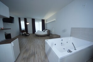 Quarto de hotel moderno com uma banheira grande e branca, uma cama redonda, uma televisão de tela plana na parede e piso de madeira.