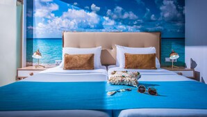 Quarto de hotel com uma cama grande, duas mesas de cabeceira, vista para o mar e uma cena de praia na parede.