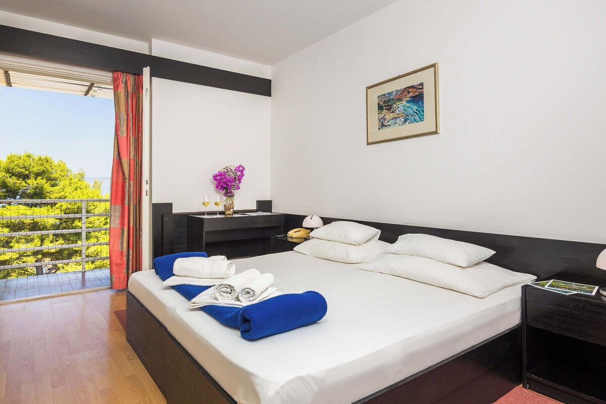 Quarto de hotel com cama, mesa de cabeceira iluminada por uma luminária, quadro na parede e vista para o verde pela janela.