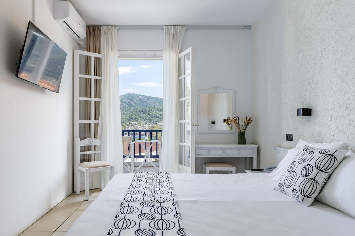 Quarto de hotel com cama, escrivaninha, cadeira e varanda com vista para as montanhas.