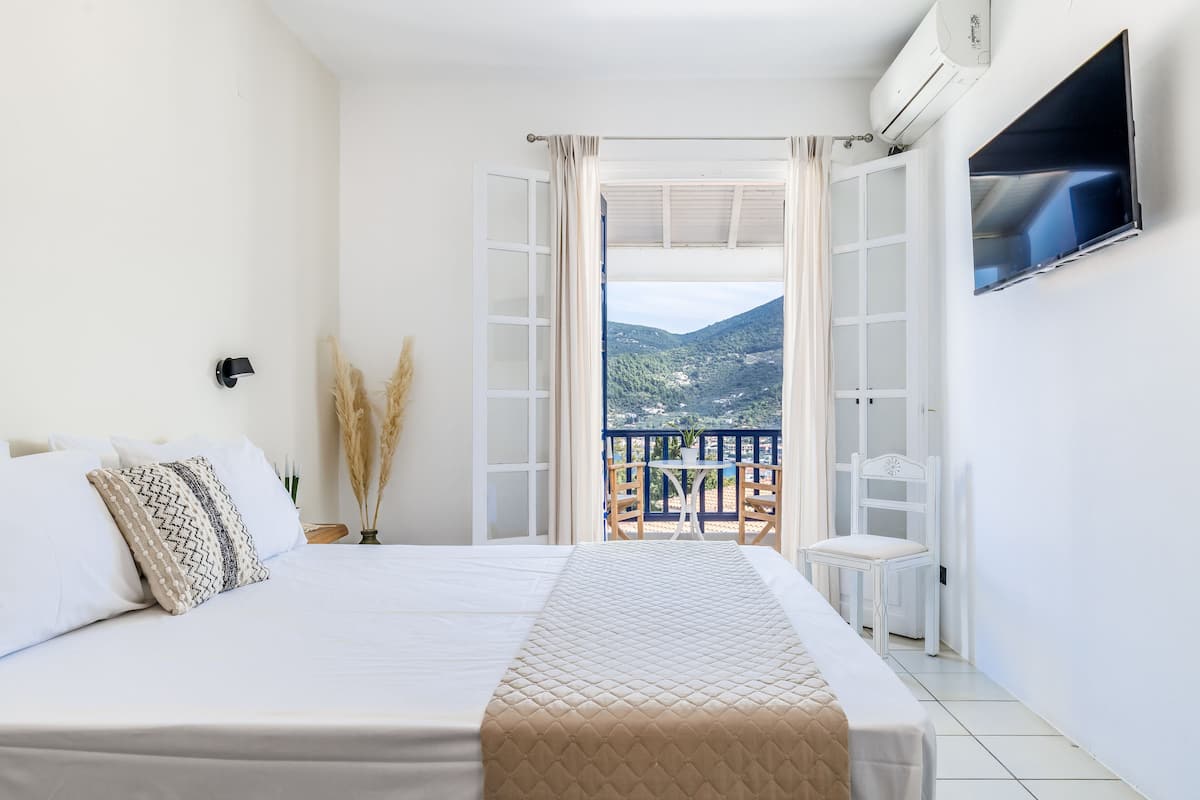 Quarto com cama, televisão, varanda com mesa e cadeira e vista para uma paisagem montanhosa.