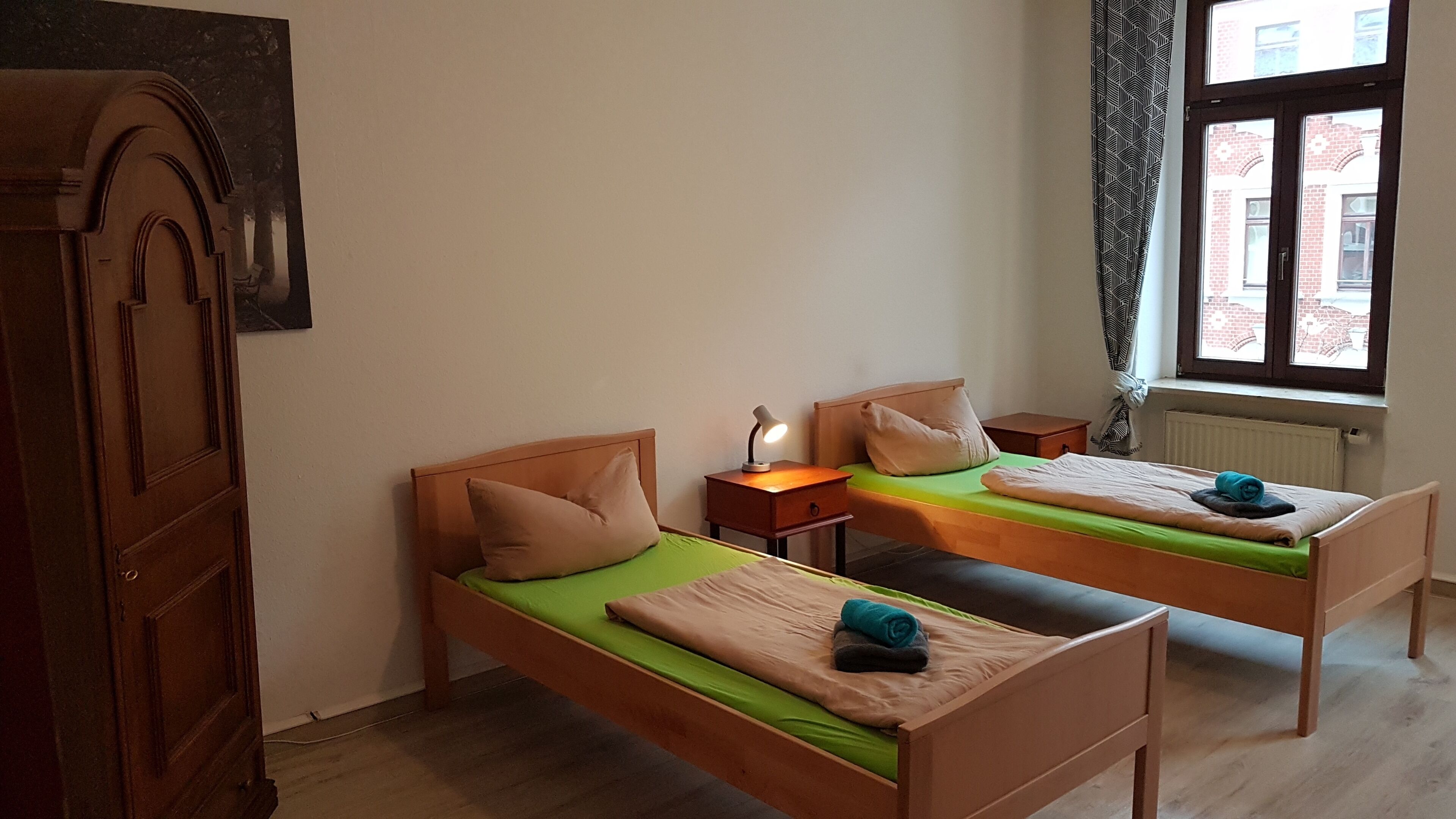 Quarto com duas camas de madeira, cada uma com colchão verde e almofadas bege. Um criado-mudo com luminária está entre as camas. Um guarda-roupa de madeira fica à esquerda, e uma janela com cortinas à direita.