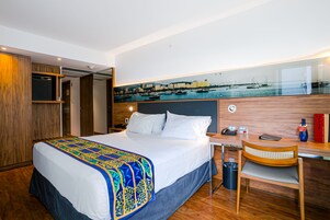 Quarto de hotel com cama, escrivaninha, cadeira, lixeira e uma grande janela com vista para a cidade.