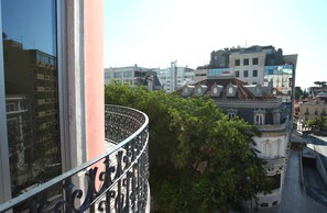 Varanda com grades de metal ornamentadas, vista para um horizonte urbano com edifícios históricos e uma rua arborizada.