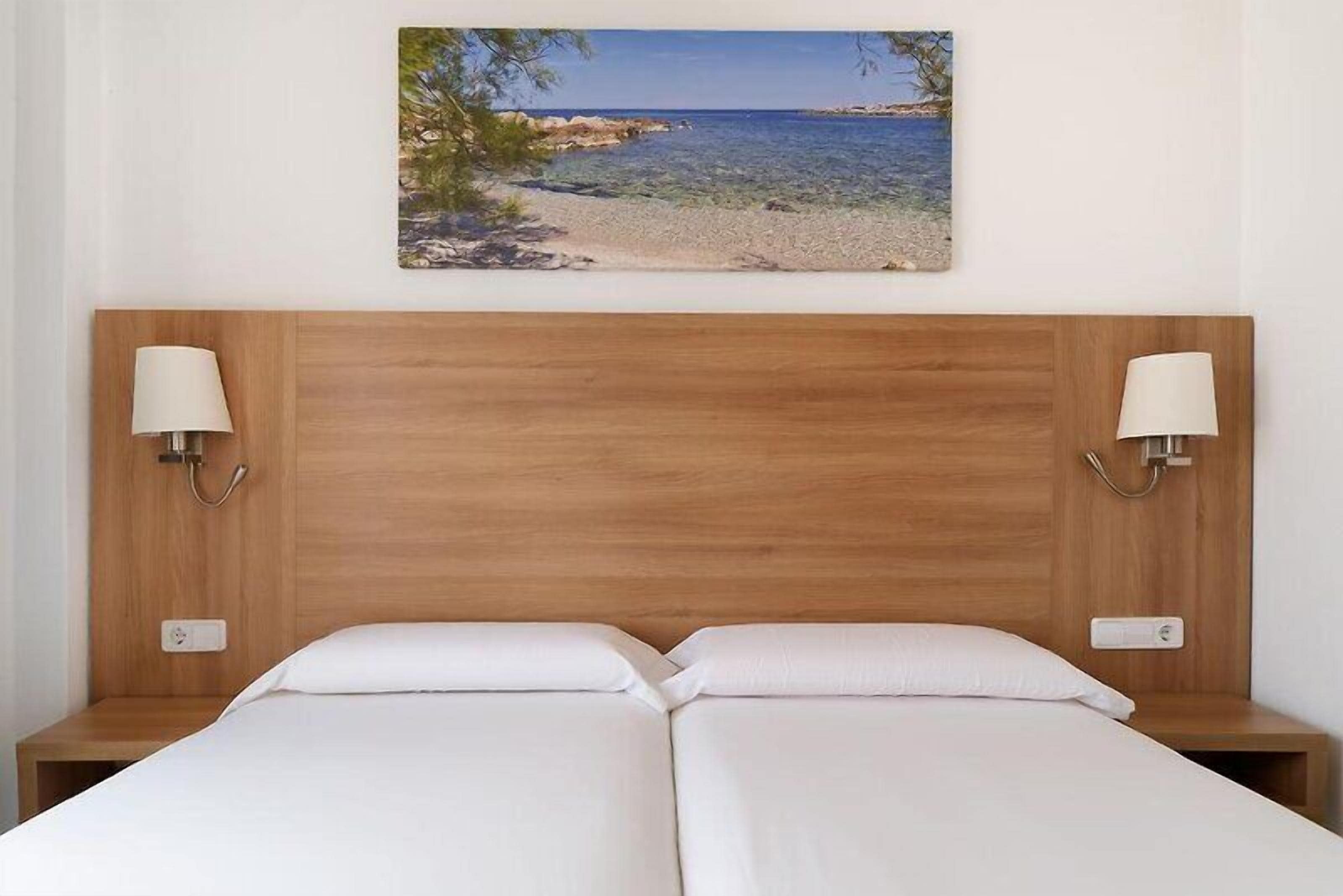 Uma cama de casal com cabeceira de madeira, duas luminárias de apoio e uma pintura de uma paisagem praiana.