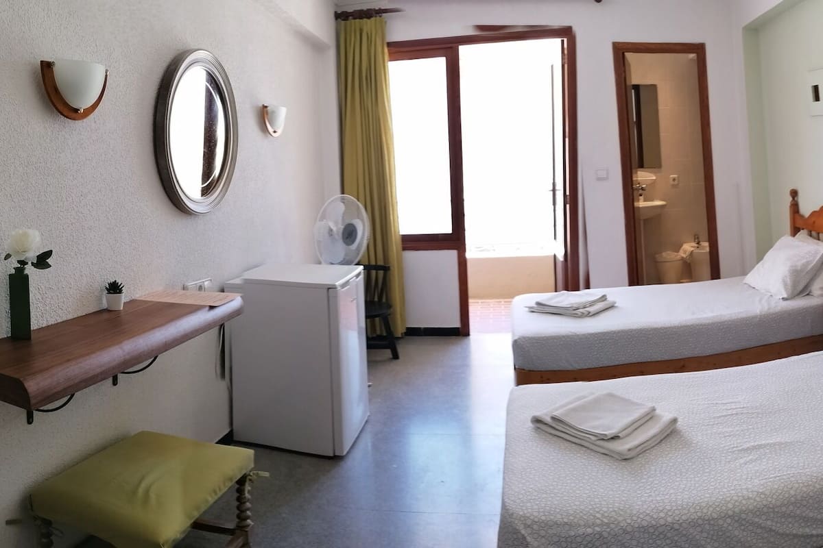 Quarto de hotel com duas camas, uma mesinha, uma cadeira, uma escrivaninha, um espelho e uma janela.