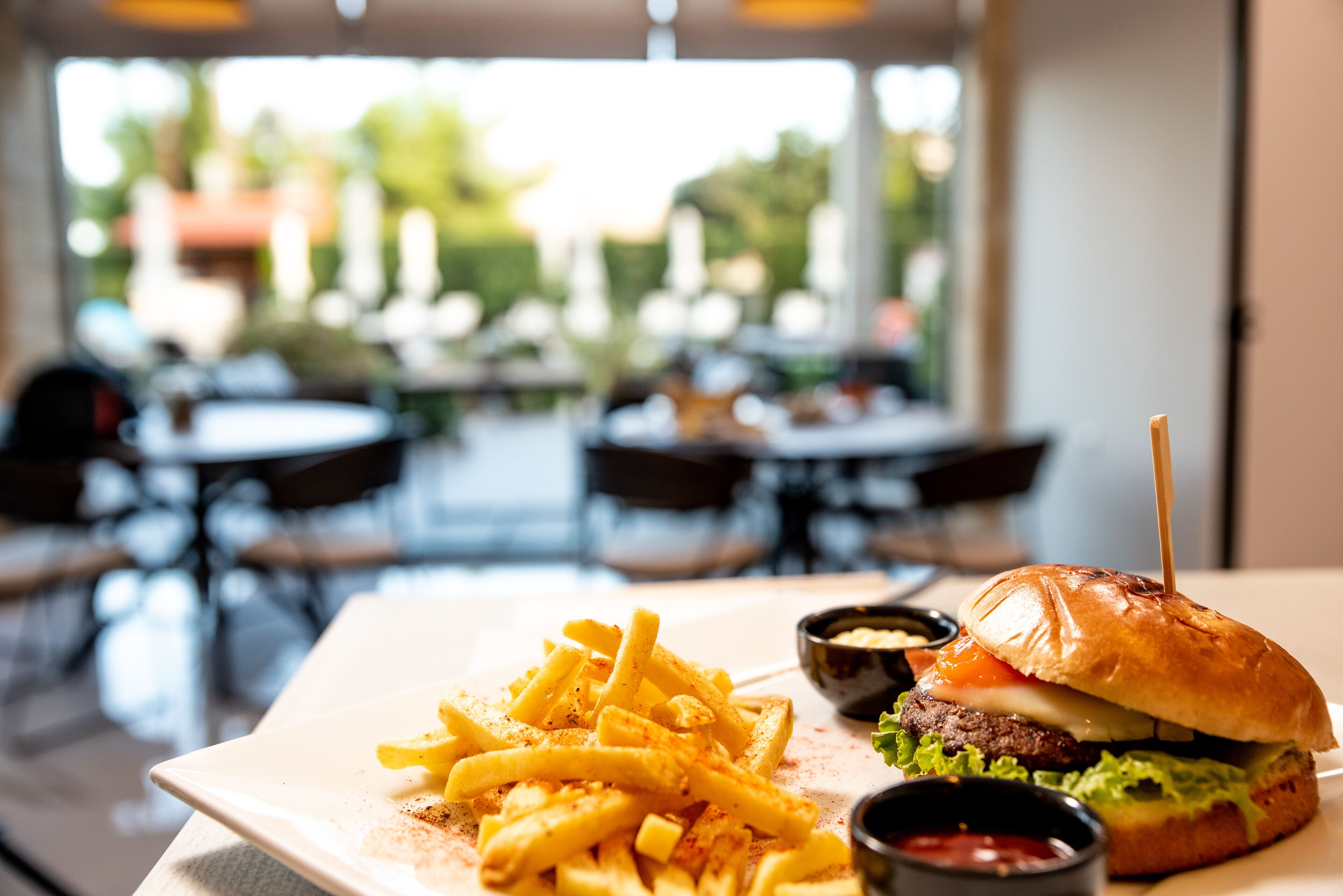 Uma refeição com um hambúrguer e batatas fritas num prato, com vista para uma área de refeições ao ar livre ao fundo.