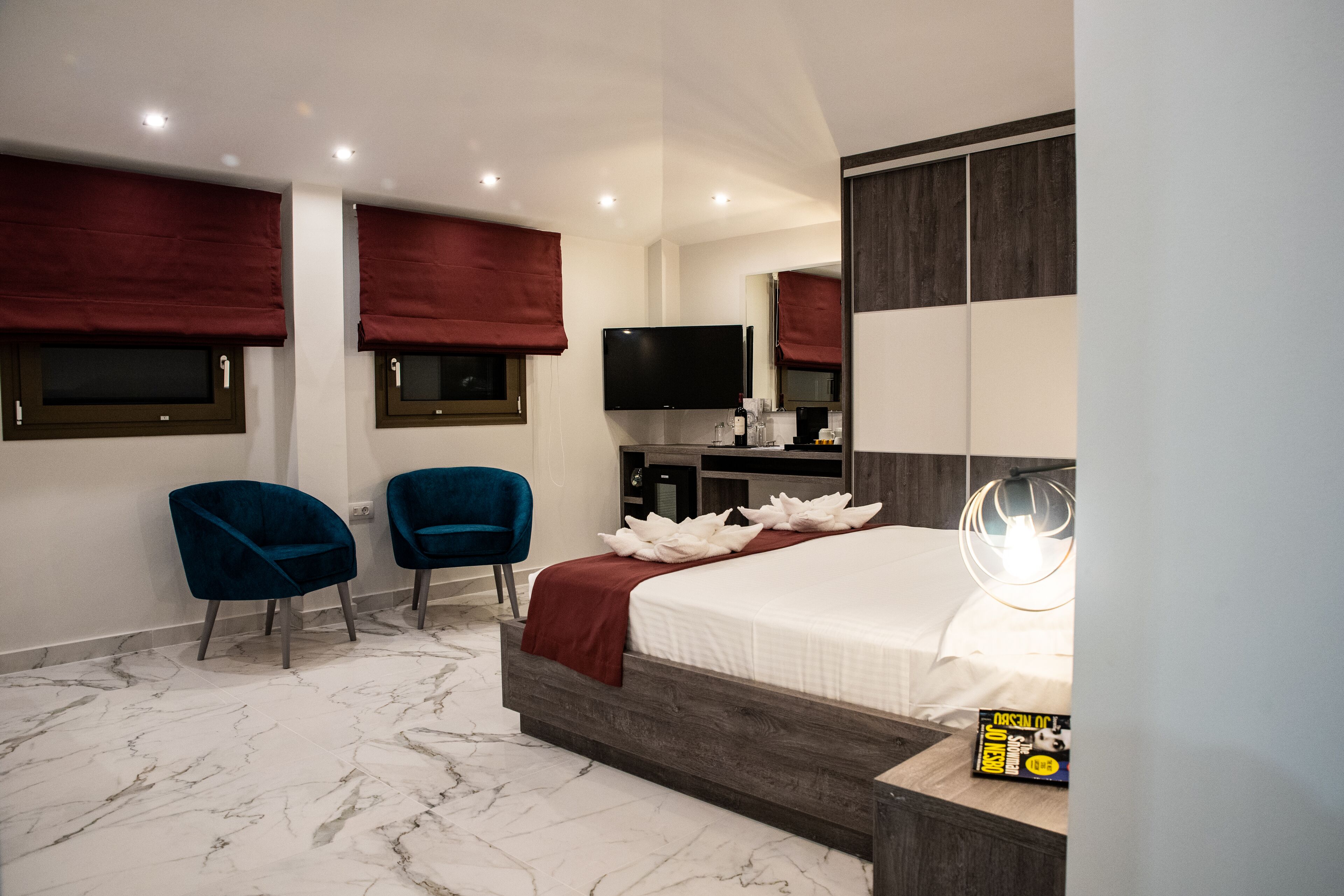 Quarto de hotel moderno com piso de mármore, cama com cabeceira de madeira, duas cadeiras azuis, escrivaninha com televisão de tela plana e um espelho grande.