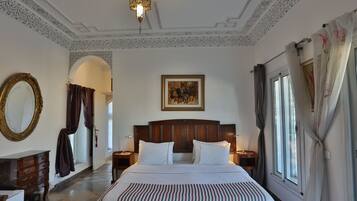 Quarto de hotel com uma cama grande, cabeceira de madeira, um lustre, um quadro emoldurado na parede e uma janela com cortinas.