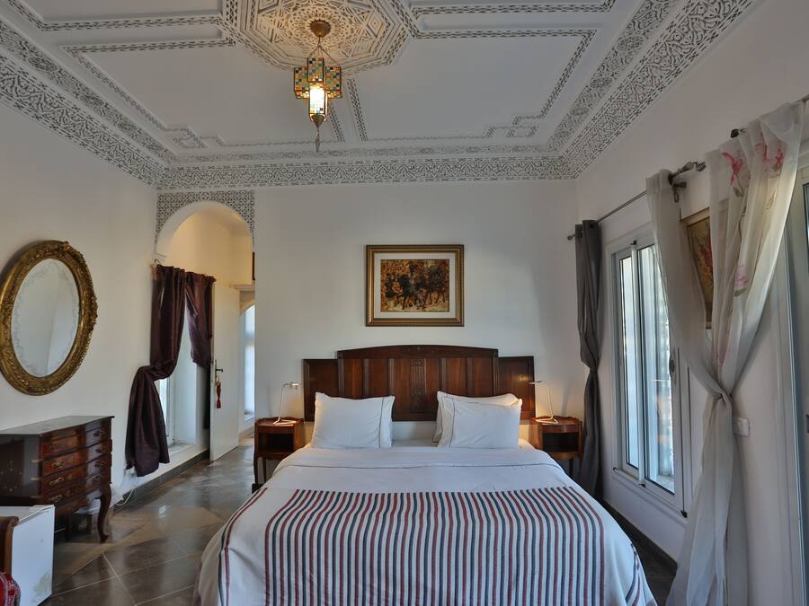 Quarto de hotel com uma cama grande, cabeceira de madeira, um lustre, um quadro emoldurado na parede e uma janela com cortinas.