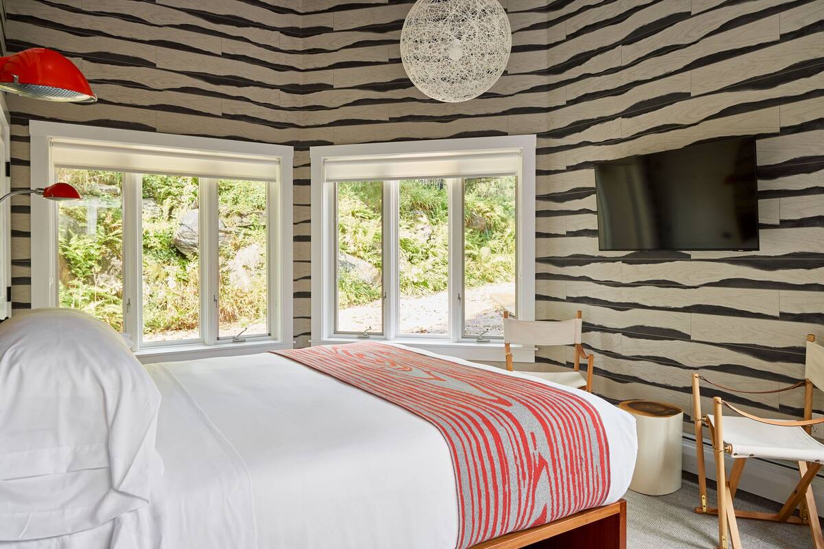Quarto com parede estampada em zebra, cama com manta listrada em vermelho e branco, luminária vermelha, luminária de teto branca e TV fixada na parede.