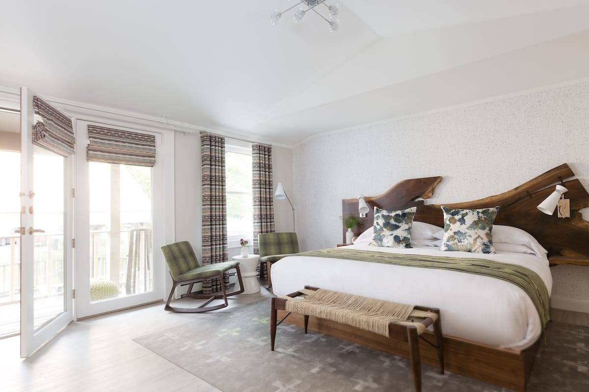 Um quarto espaçoso com uma cama grande, cabeceira de madeira, um banquinho, duas cadeiras e vista para o exterior através de uma porta de vidro deslizante.