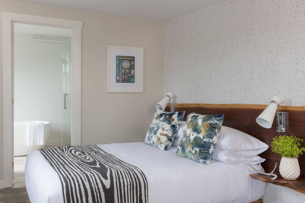 Uma cama bem arrumada com colcha estampada, duas almofadas decorativas, um abajur e um pequeno vaso de planta sobre uma mesinha de cabeceira de madeira.