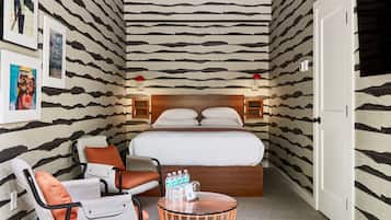 Quarto de hotel pequeno com parede listrada em preto e branco, cama com lençóis brancos, duas poltronas com almofadas laranja, mesa de centro de vidro com garrafas de água e duas luminárias de parede para leitura.