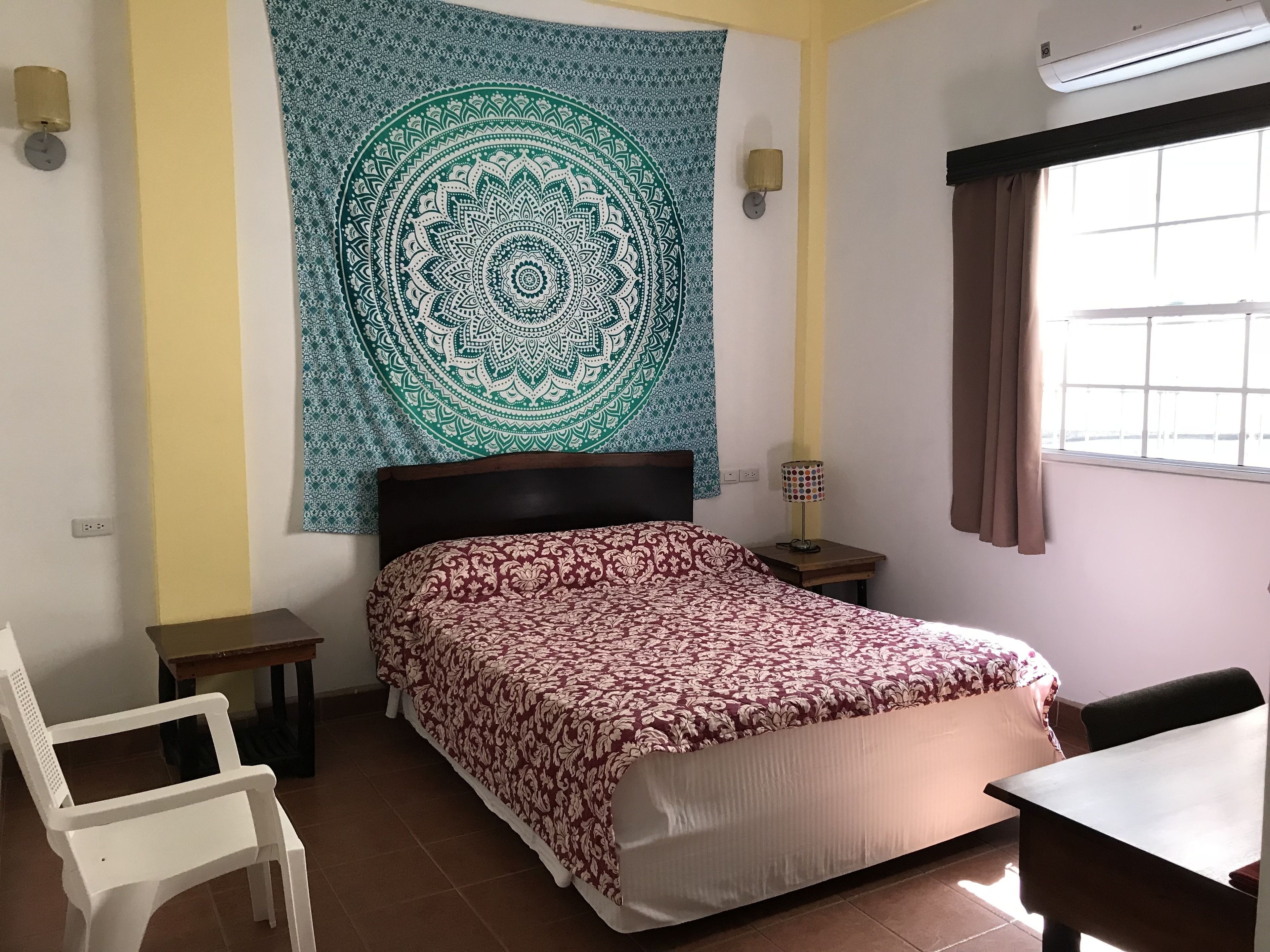 Quarto com cama, cadeira, mesa pequena, tapeçaria e janela com cortinas.