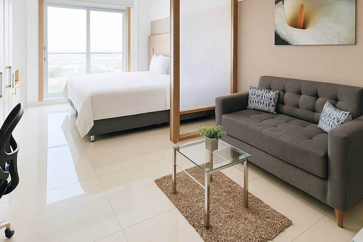 Quarto de hotel moderno com cama, sofá, mesa de centro de vidro e uma janela grande.