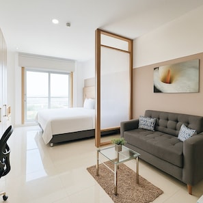 Quarto de hotel moderno com cama, sofá, mesa de centro de vidro e uma janela grande.