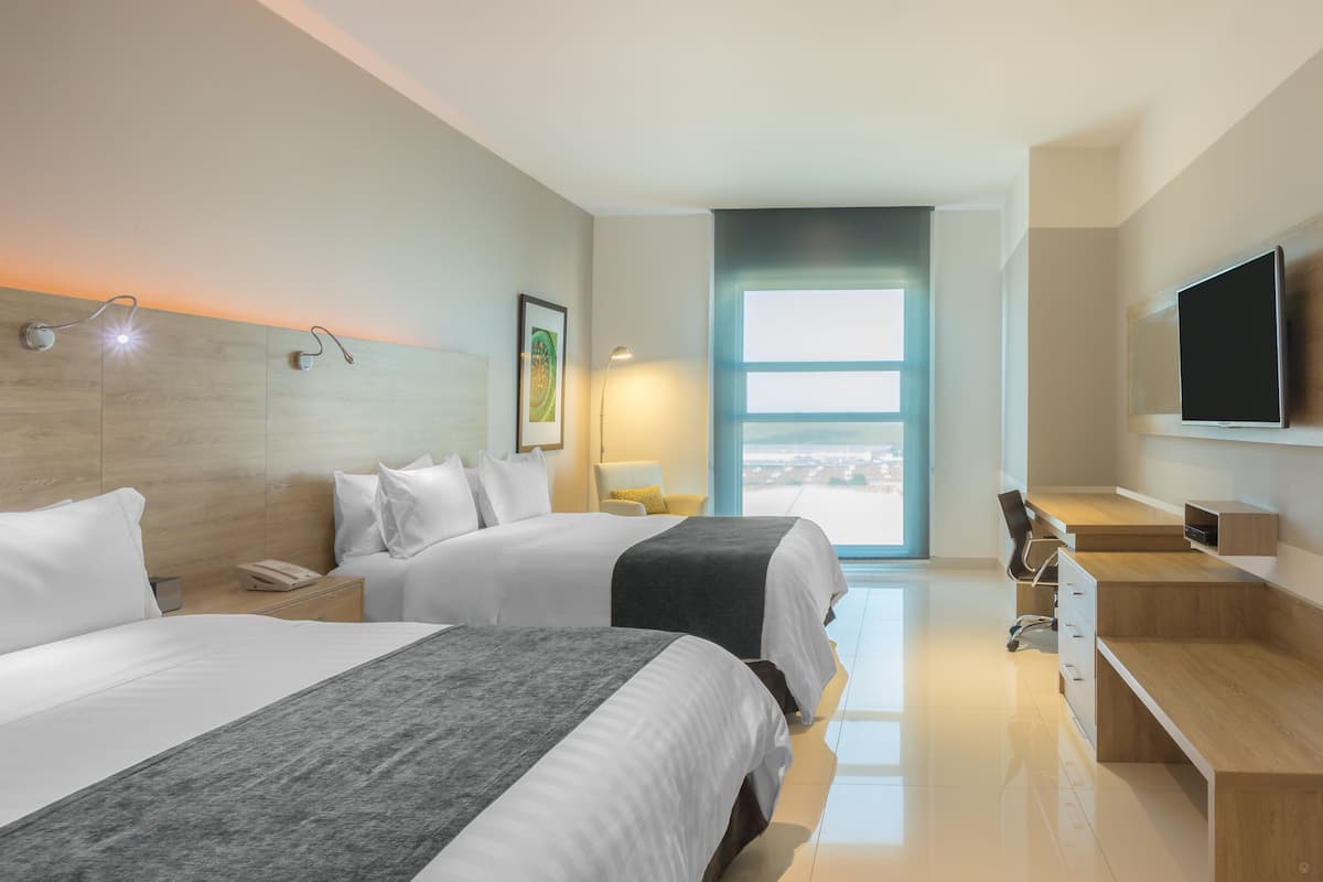 Quarto de hotel com duas camas, uma escrivaninha, uma cadeira, uma TV e uma janela grande.