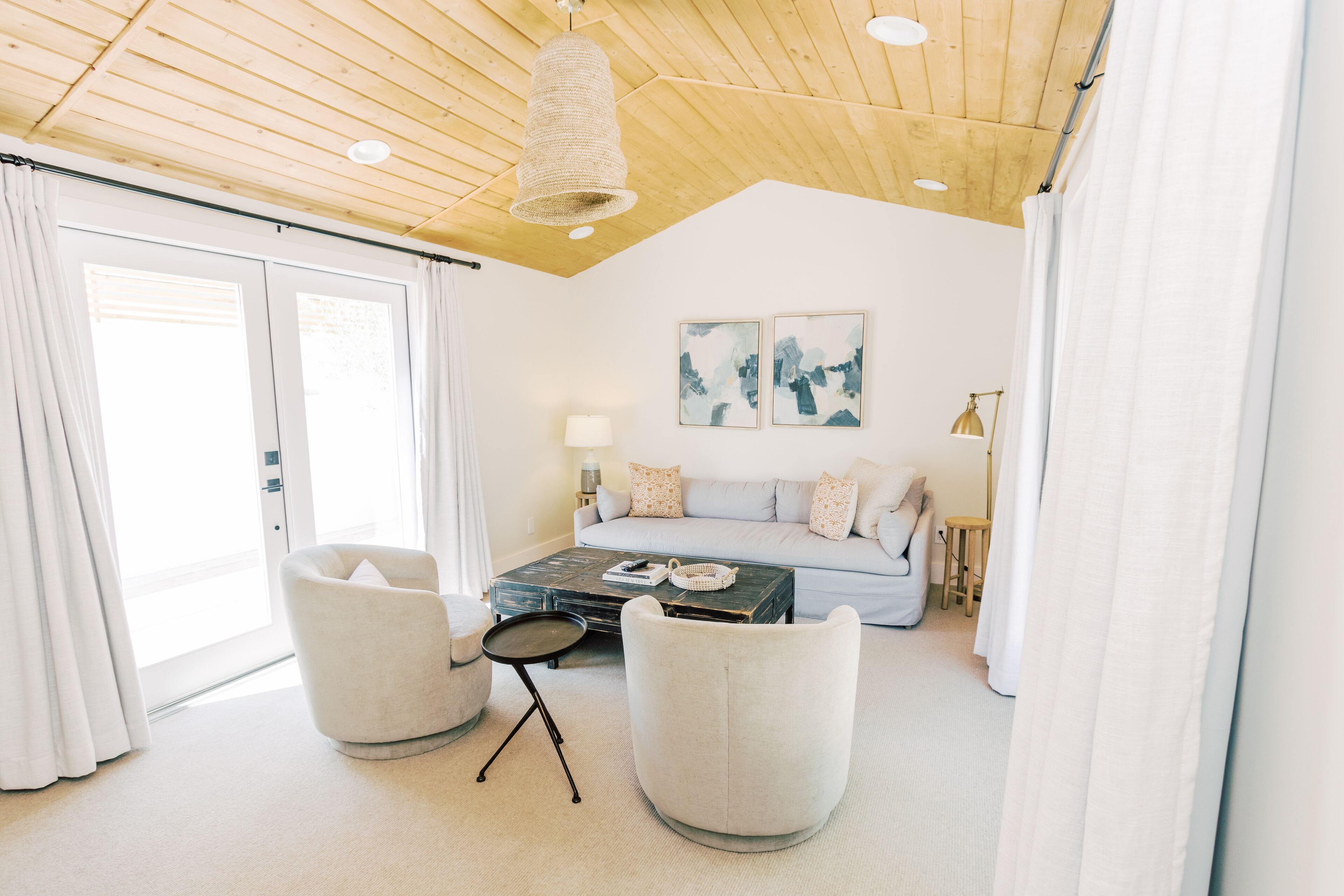 Uma sala de estar moderna com teto de madeira, um sofá, duas poltronas, uma mesa de centro e uma janela grande com cortinas.