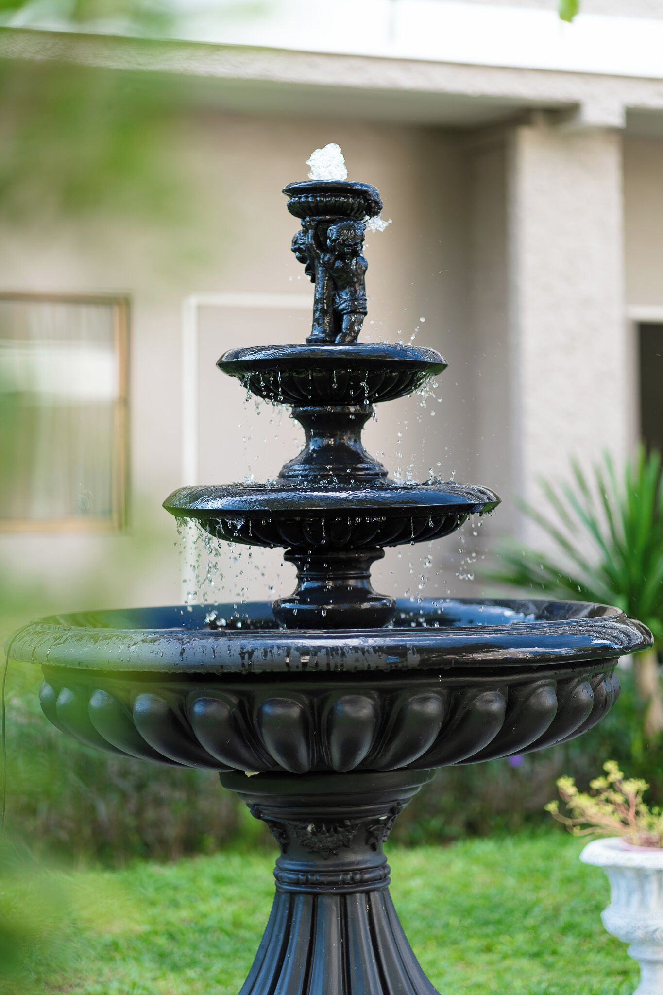 Uma fonte de vários níveis com água jorrando do nível superior, situada num jardim com um edifício ao fundo.