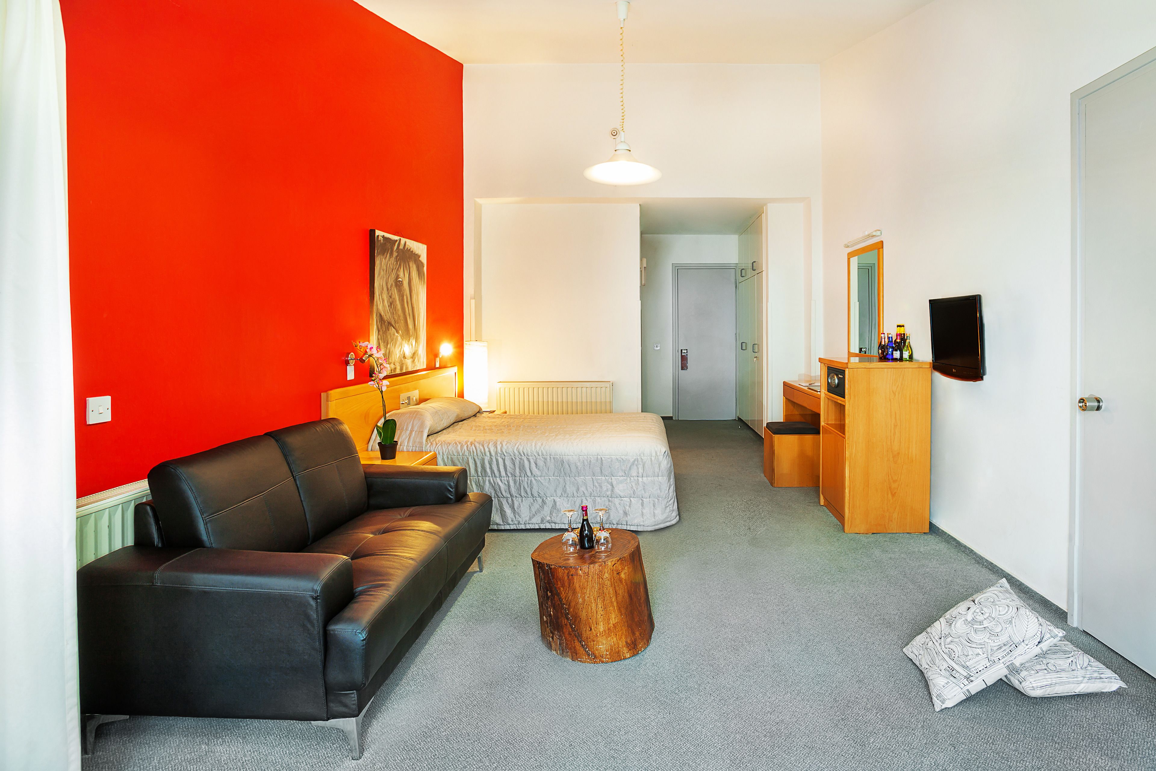 Quarto de hotel com parede vermelha de destaque, sofá de couro preto, cama com colcha branca, cómoda de madeira e mesa de centro redonda de madeira.