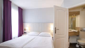 Quarto de hotel com cama, mesa de cabeceira, luminária na parede, banheiro com vaso sanitário e chuveiro.