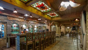 Uma área de bar com cadeiras de madeira e um balcão de pedra, apresentando janelas de vitrais e vigas de madeira no teto.