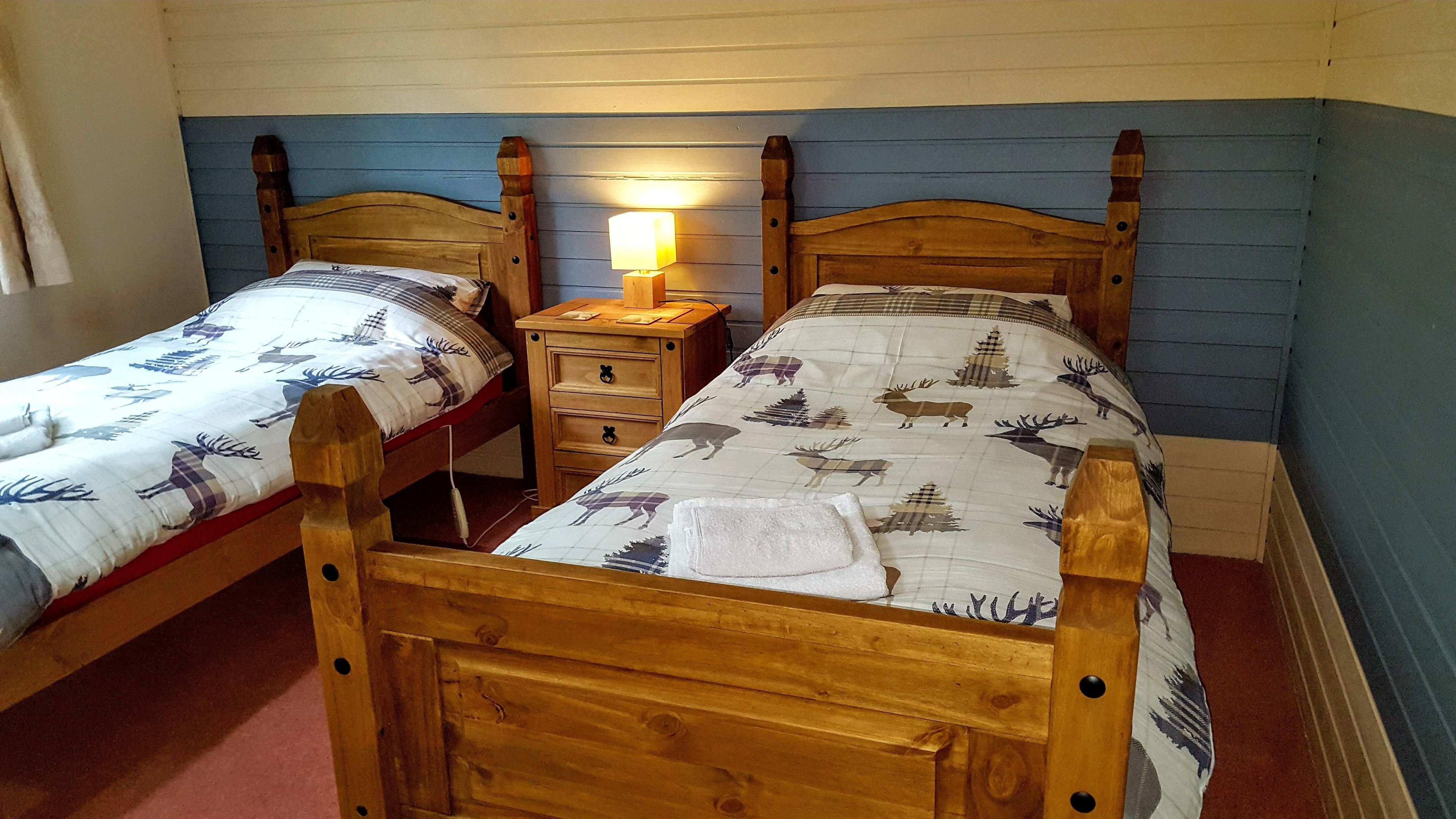 Duas camas de madeira com cabeceiras e peseiras iguais, uma mesa de cabeceira com abajur e uma janela com venezianas horizontais.