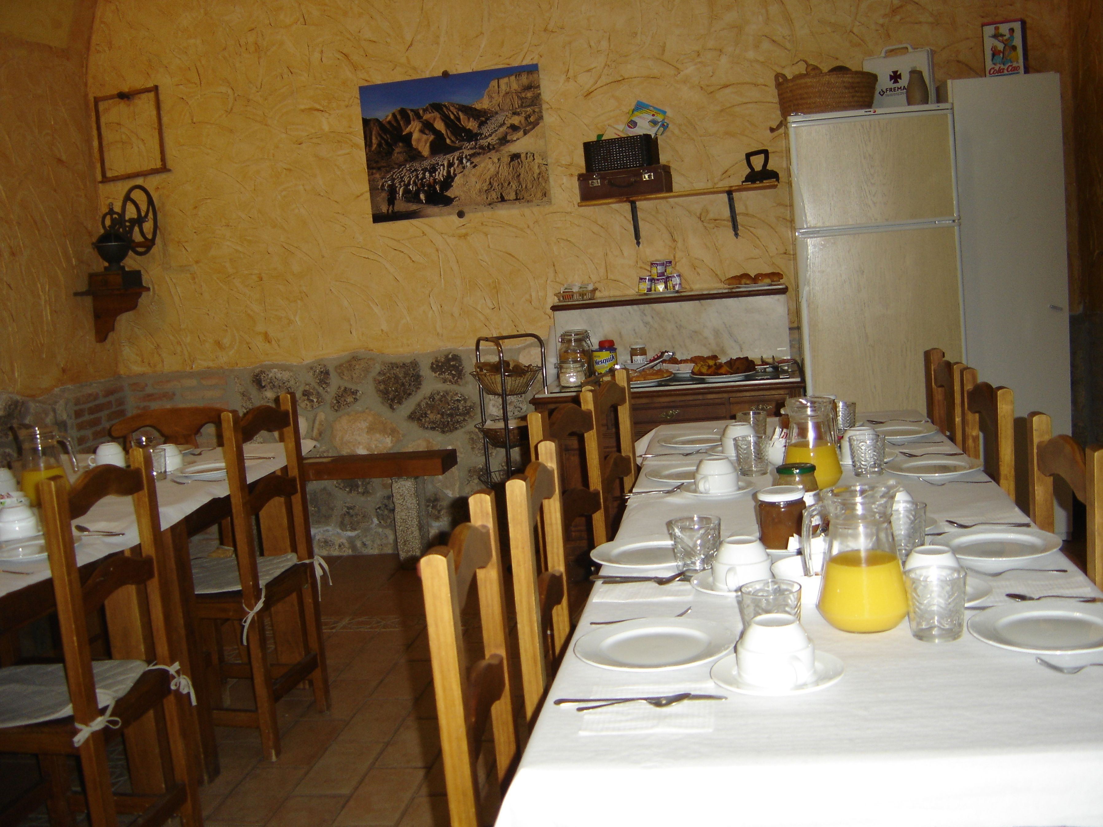 Uma sala de jantar com uma mesa comprida posta para refeição, cadeiras de madeira, uma parede de pedra e uma geladeira ao fundo.