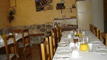 Uma sala de jantar com uma mesa comprida posta para refeição, cadeiras de madeira, uma parede de pedra e uma geladeira ao fundo.