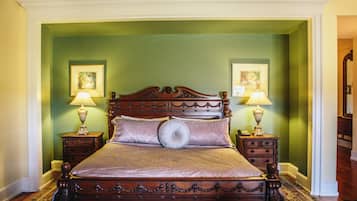 Uma cama de madeira grande e ornamentada, com cabeceira combinando, flanqueada por duas mesas de cabeceira com abajures, apoiada em uma parede verde com obras de arte emolduradas.