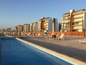 Uma área à beira da piscina com espreguiçadeiras e piscina, cercada por edifícios modernos com sacadas.